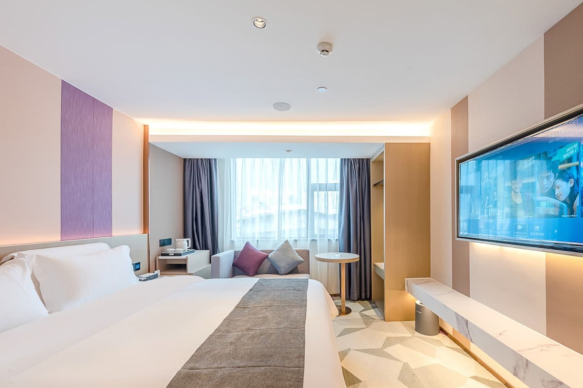 Quarto de hotel com uma cama grande, televisão na parede, uma secretária pequena e uma área de estar com sofá e poltrona.
