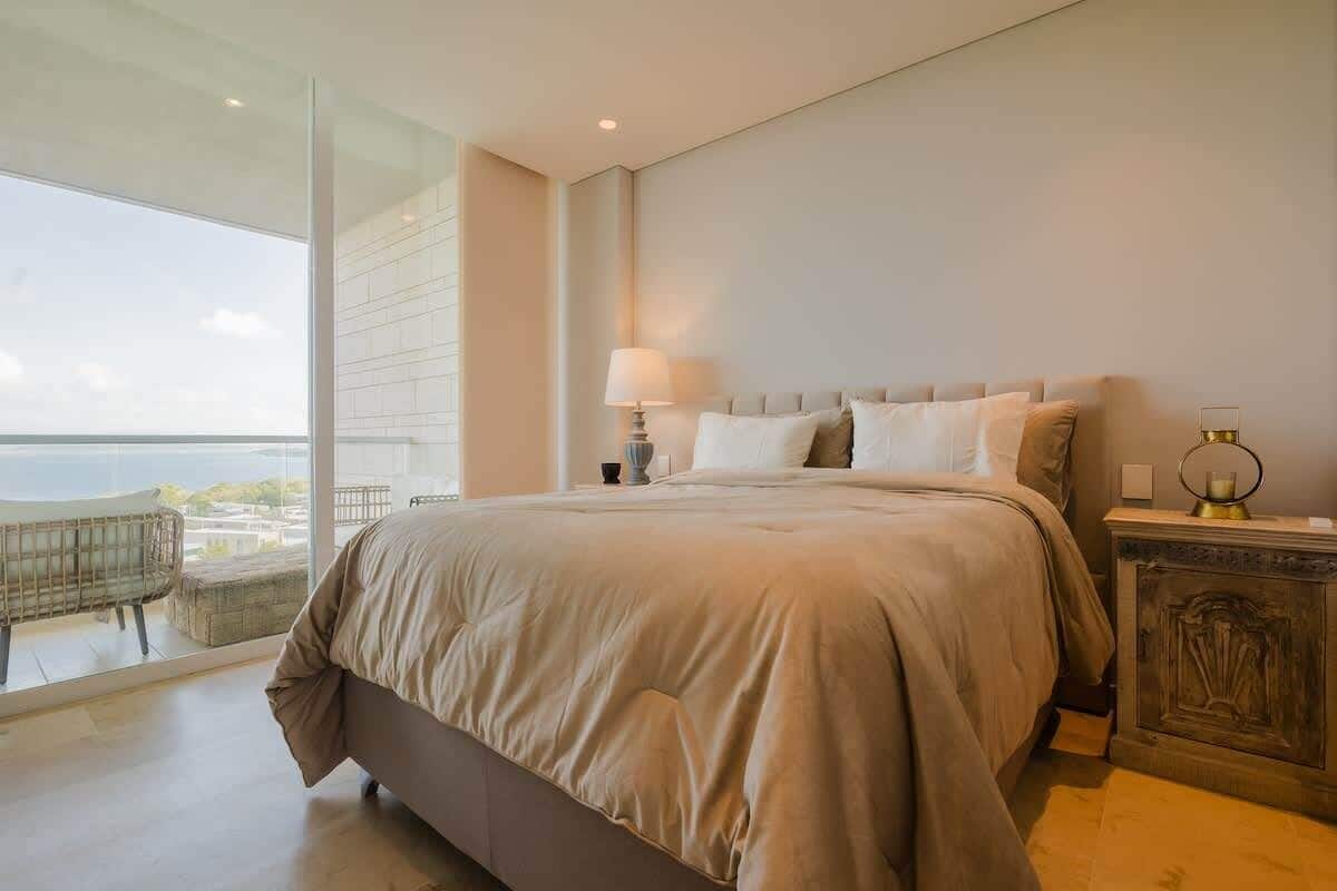 Quarto com cama grande, mesa de cabeceira, luminária e vista para o exterior.