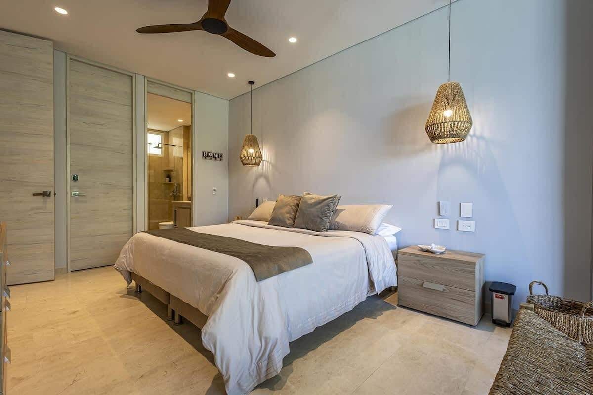 Quarto moderno com uma cama grande, uma mesinha de cabeceira de madeira, uma cesta trançada e um ventilador de teto.