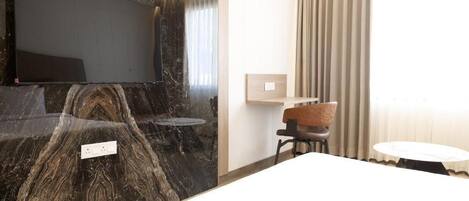 Quarto de hotel moderno com uma televisão de tela plana grande fixada em uma parede de mármore, uma cadeira de madeira, uma mesa redonda pequena e uma cama com cabeceira branca.