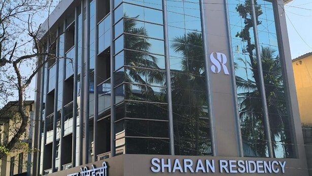 Um edifício moderno com a placa 'SHARAN RESIDENCY' e um logotipo com 'SR'.