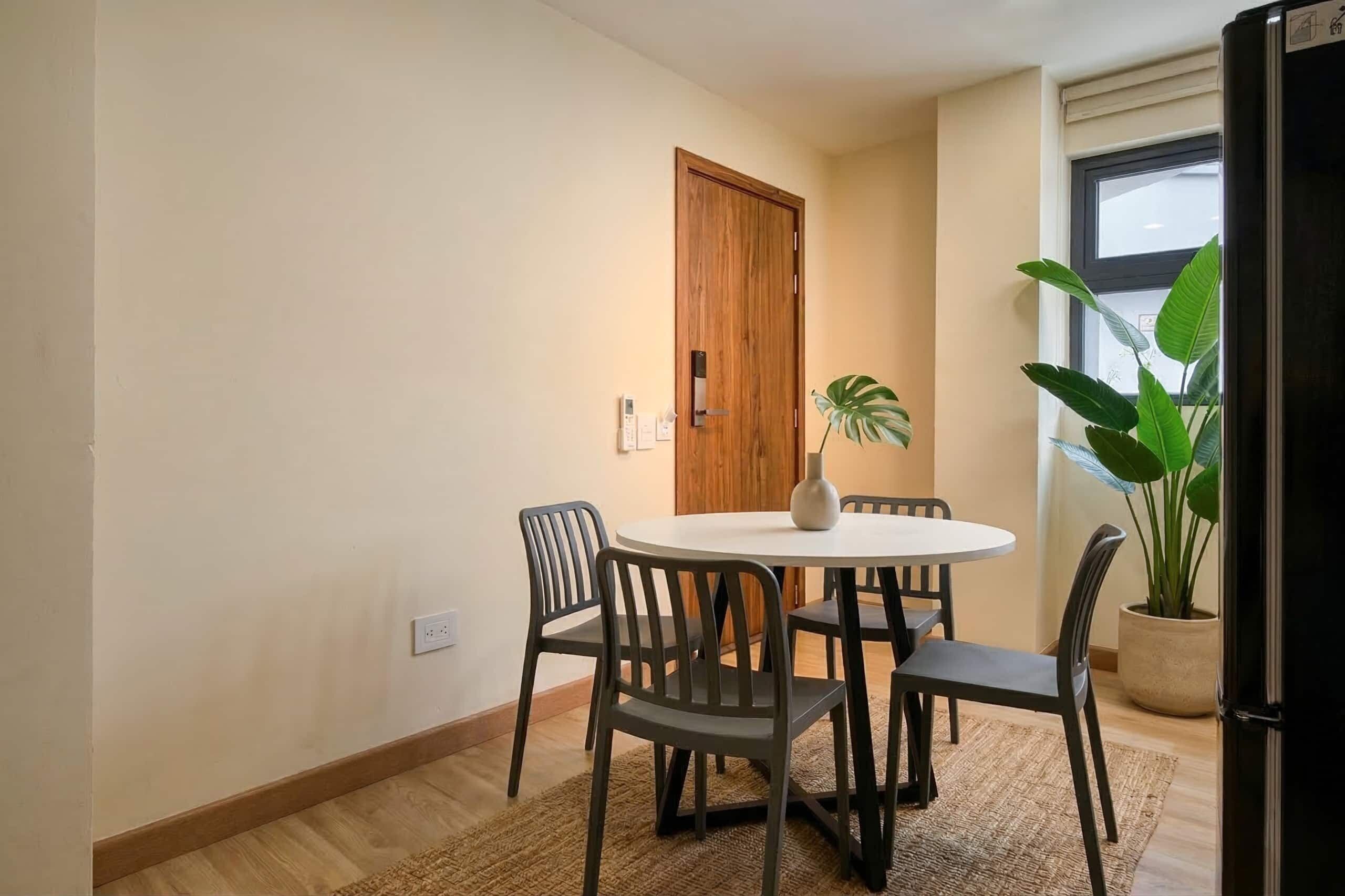 Uma sala de jantar com uma mesa redonda, quatro cadeiras, um vaso de planta e uma porta de madeira.