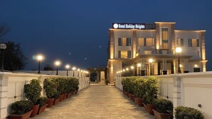 Entrada de hotel com caminho de paralelepípedos, plantas em vasos e uma placa com a inscrição 'Hotel Holiday Heights'.