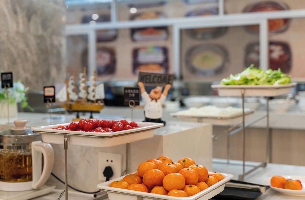 Uma área de buffet com diversos alimentos, incluindo frutas e vegetais, e um letreiro de boas-vindas.