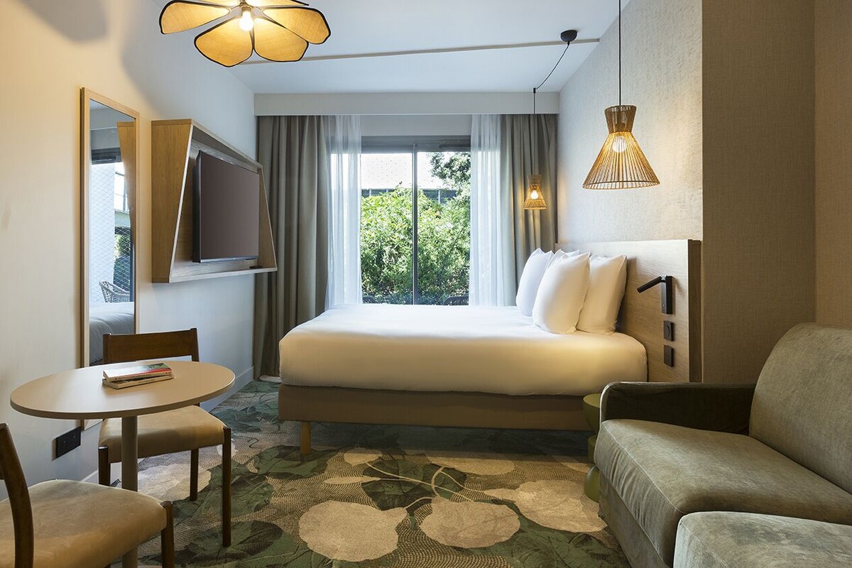 Quarto de hotel com uma cama, um sofá, uma televisão e uma janela com cortinas.