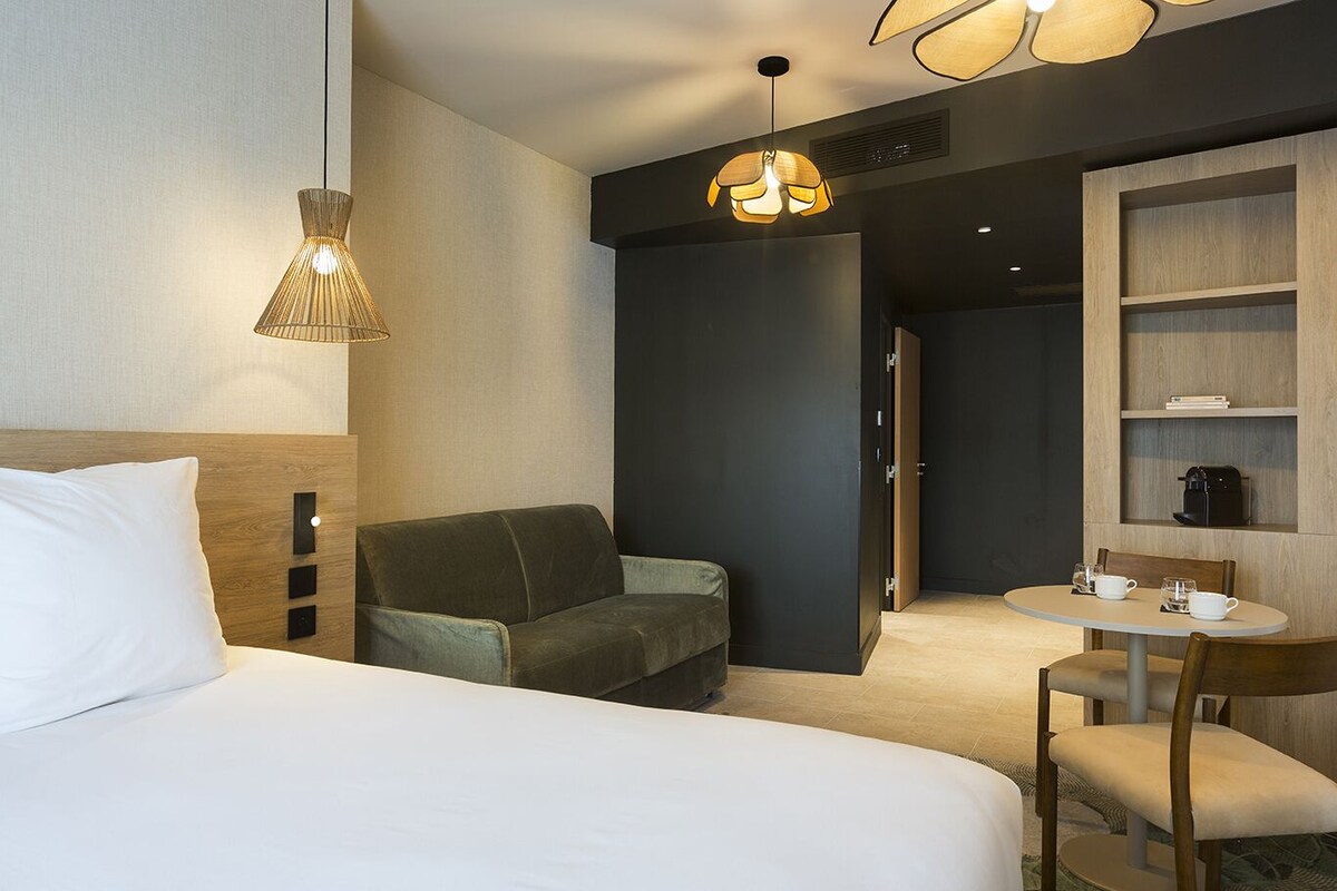 Quarto de hotel com cama, poltrona verde, mesa redonda com cadeiras e prateleira embutida com máquina de café.