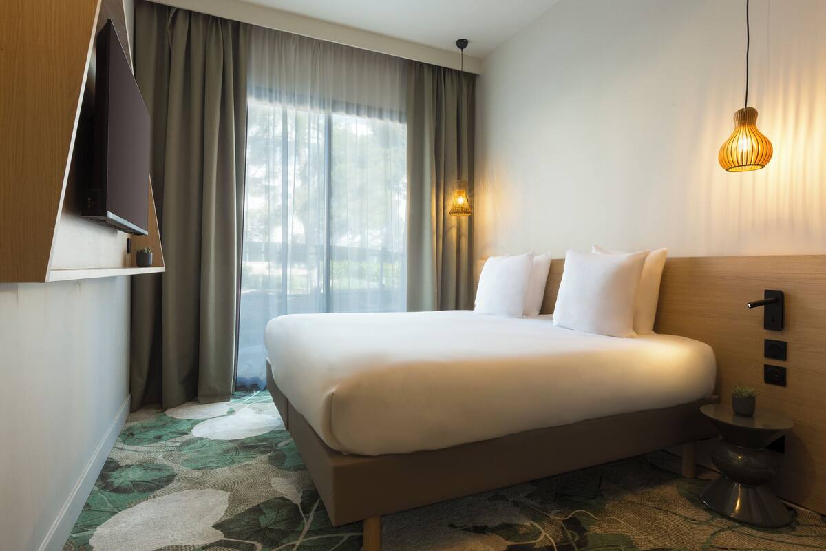 Quarto de hotel com cama, televisão, pequena mesa com planta, luminária na parede e janela com cortinas.
