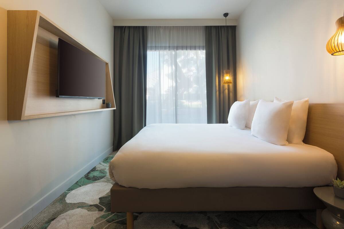Quarto de hotel com cama, televisão na parede, luminária de cabeceira e janela com cortinas.