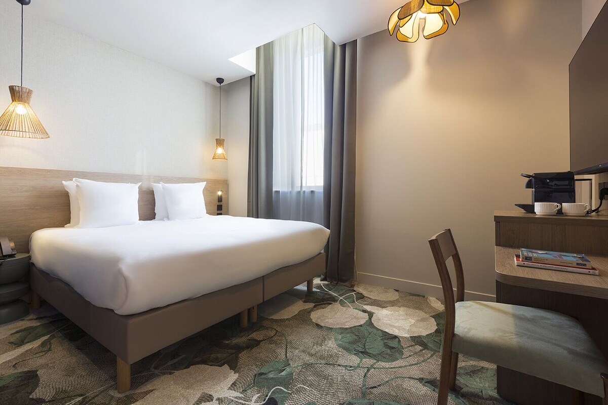Quarto de hotel com cama, mesa de cabeceira, abajur, cadeira e escrivaninha com cafeteira.