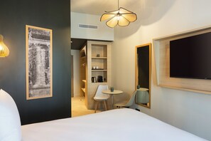 Quarto de hotel com cama, escrivaninha com cadeiras, televisão, quadro na parede e luminária de teto.