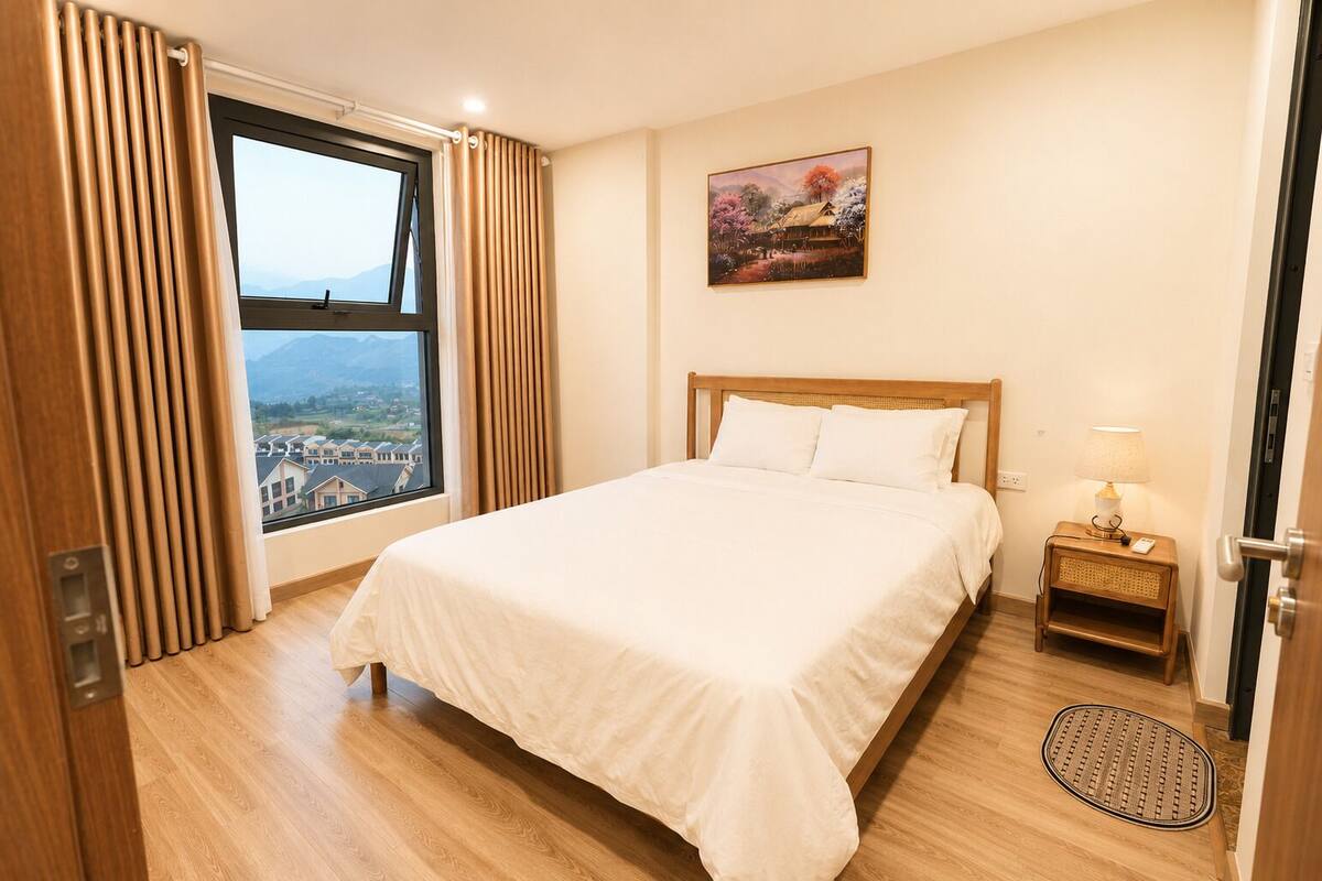 Quarto de hotel com uma cama grande, uma mesa de cabeceira com abajur, um quadro na parede e uma janela com cortinas.