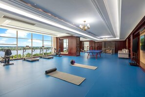 Quarto espaçoso com equipamentos de ginástica, mesa de pingue-pongue e grandes janelas com vista para um corpo de água.