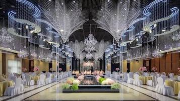 Um salão de banquetes com palco central, rodeado por mesas e cadeiras, e teto decorado com lustres e luzes pendentes.