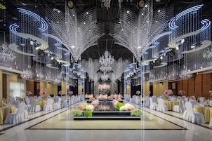 Um salão de banquetes com palco central, rodeado por mesas e cadeiras, e teto decorado com lustres e luzes pendentes.
