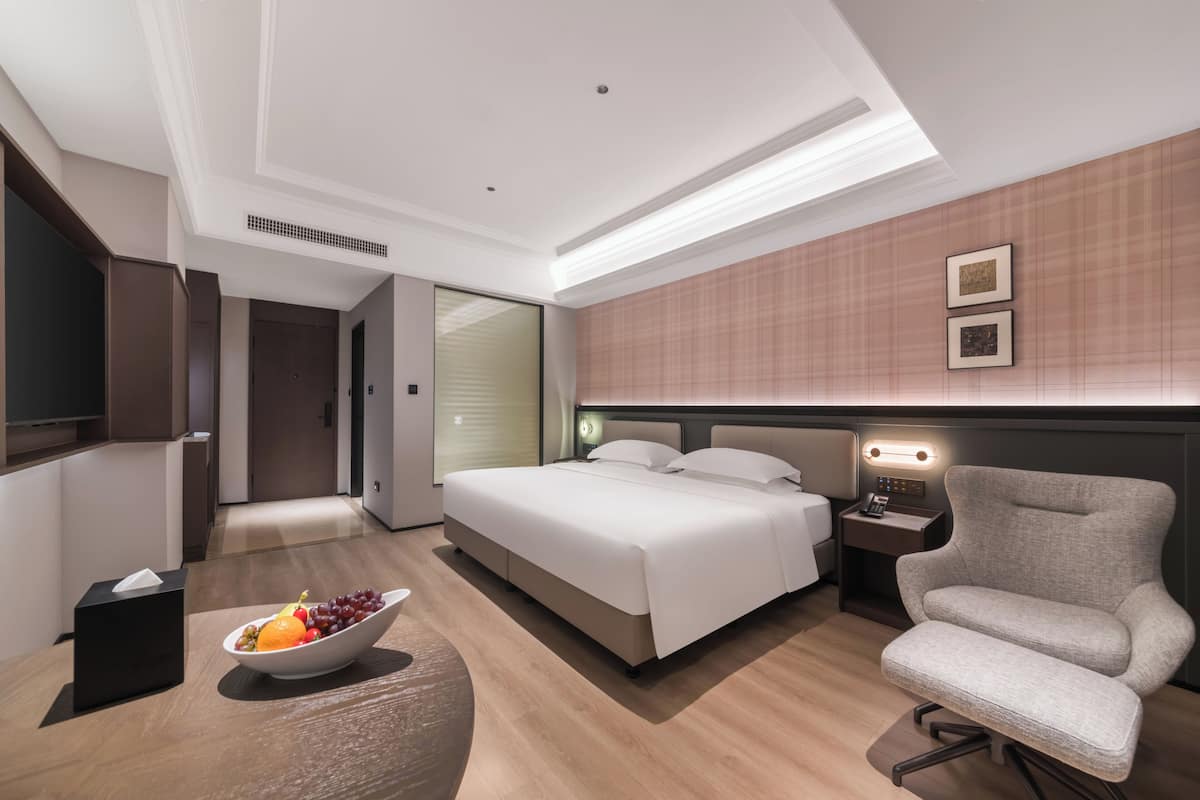 Quarto de hotel moderno com uma cama grande, área de estar, mesa de cabeceira com abajur e televisão na parede.