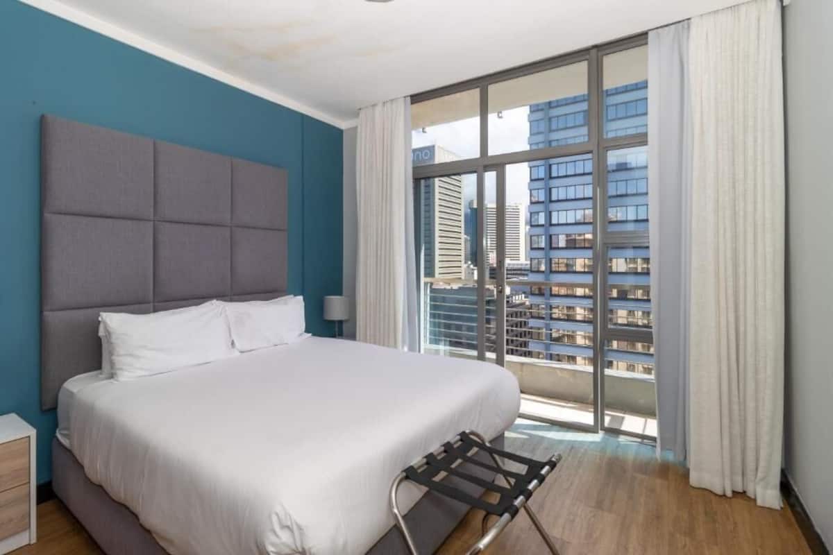 Quarto de hotel com uma cama grande, cabeceira, mesa de cabeceira, janela com vista para edifícios e porta deslizante.