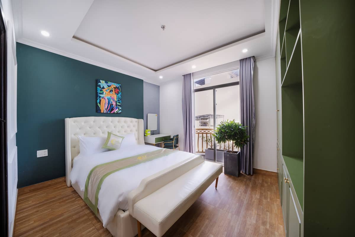 Quarto de hotel moderno com uma cama grande, uma mesa de escritório, uma planta em vaso e uma varanda com cortinas.