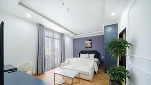 Quarto de hotel com cama, sofá, mesa de centro, televisão, planta e janela com cortinas.