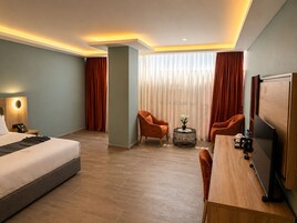 Quarto de hotel com cama, duas poltronas, escrivaninha com computador, mesinha com um vaso e uma janela grande com cortinas.