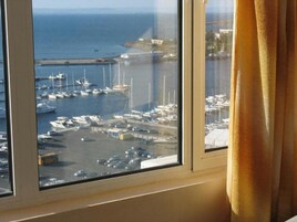 Vista pela janela com cortina, mostrando uma marina com barcos e a costa ao longe.