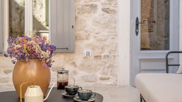 Uma parede de pedra, uma cama, uma mesa com um vaso de flores e uma cafeteira.