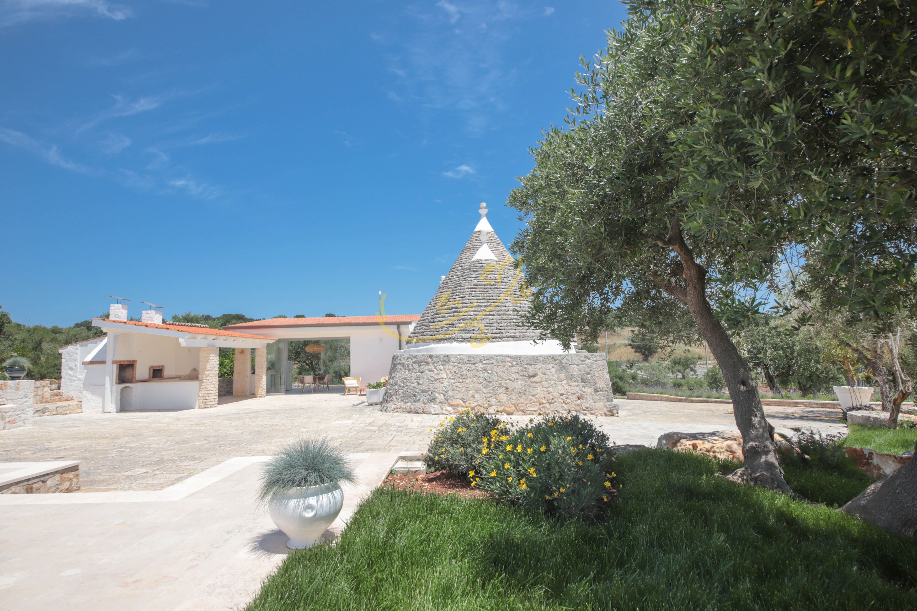 Uma casa moderna com uma pirâmide de pedra, um alpendre coberto e um jardim bem cuidado, com uma árvore grande e flores amarelas.