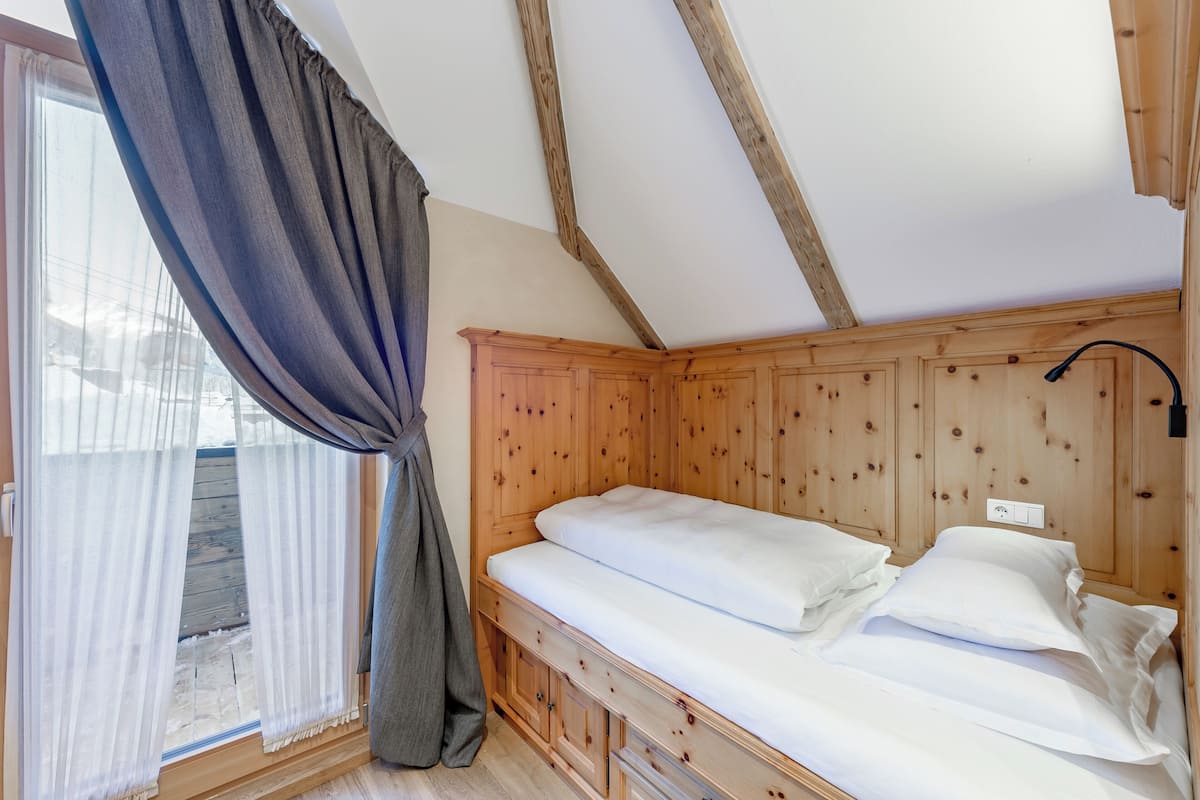 Quarto de cabana de madeira com uma cama, janela com cortinas e teto com vigas aparentes.