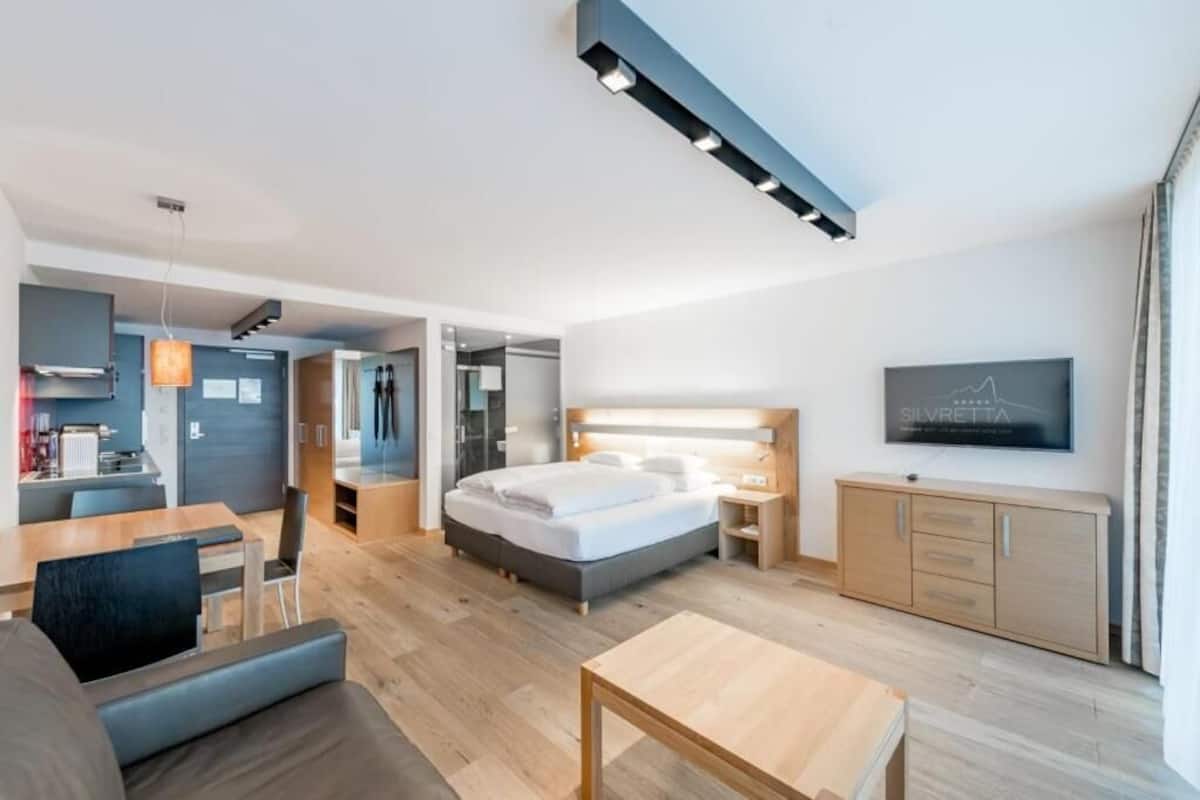Quarto de hotel moderno com cama, televisão, escrivaninha, sofá e área de refeições.
