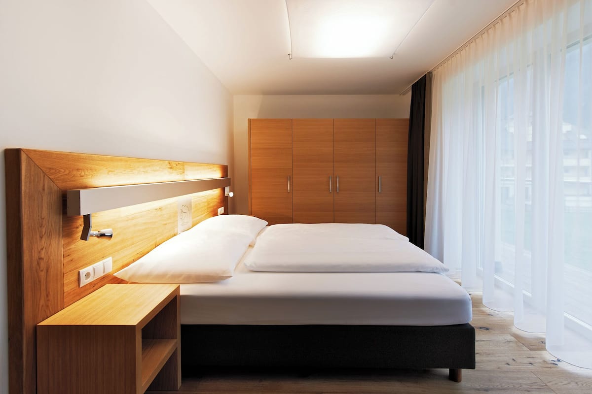 Quarto moderno com cabeceira de madeira, mesa de cabeceira, cama com roupa de cama branca e um guarda-roupa grande.