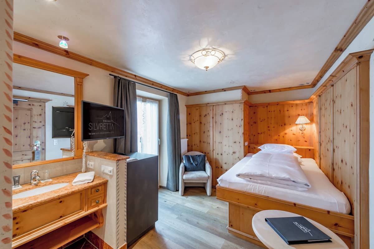 Quarto de hotel com decoração em madeira, uma cama, uma escrivaninha com computador, uma cadeira e uma televisão.