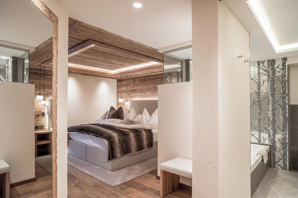 Quarto moderno com teto de madeira, cama com roupa de cama branca, mesa de cabeceira e banheiro com chuveiro revestido em azulejos.