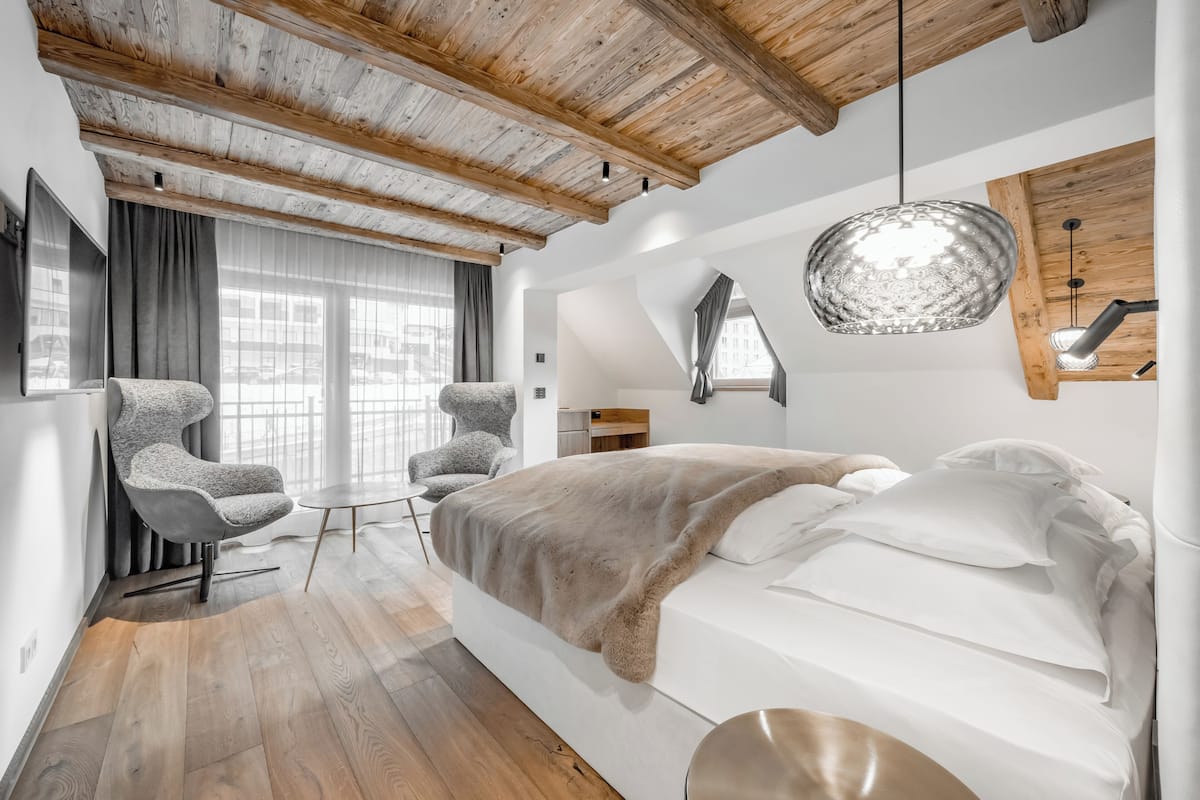 Quarto moderno com teto de madeira, uma cama grande, duas cadeiras, uma mesa redonda e um lustre.