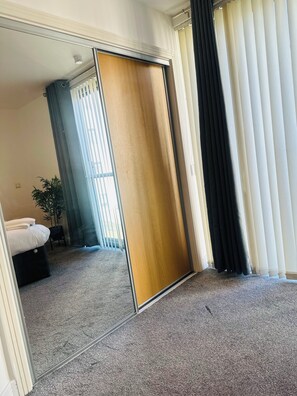 Quarto de hotel com um espelho grande, uma cama, uma planta e uma janela com persianas.