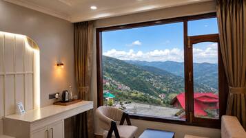 Quarto de hotel com cama, escrivaninha, cadeira, mesa pequena e uma grande janela com vista para as montanhas.