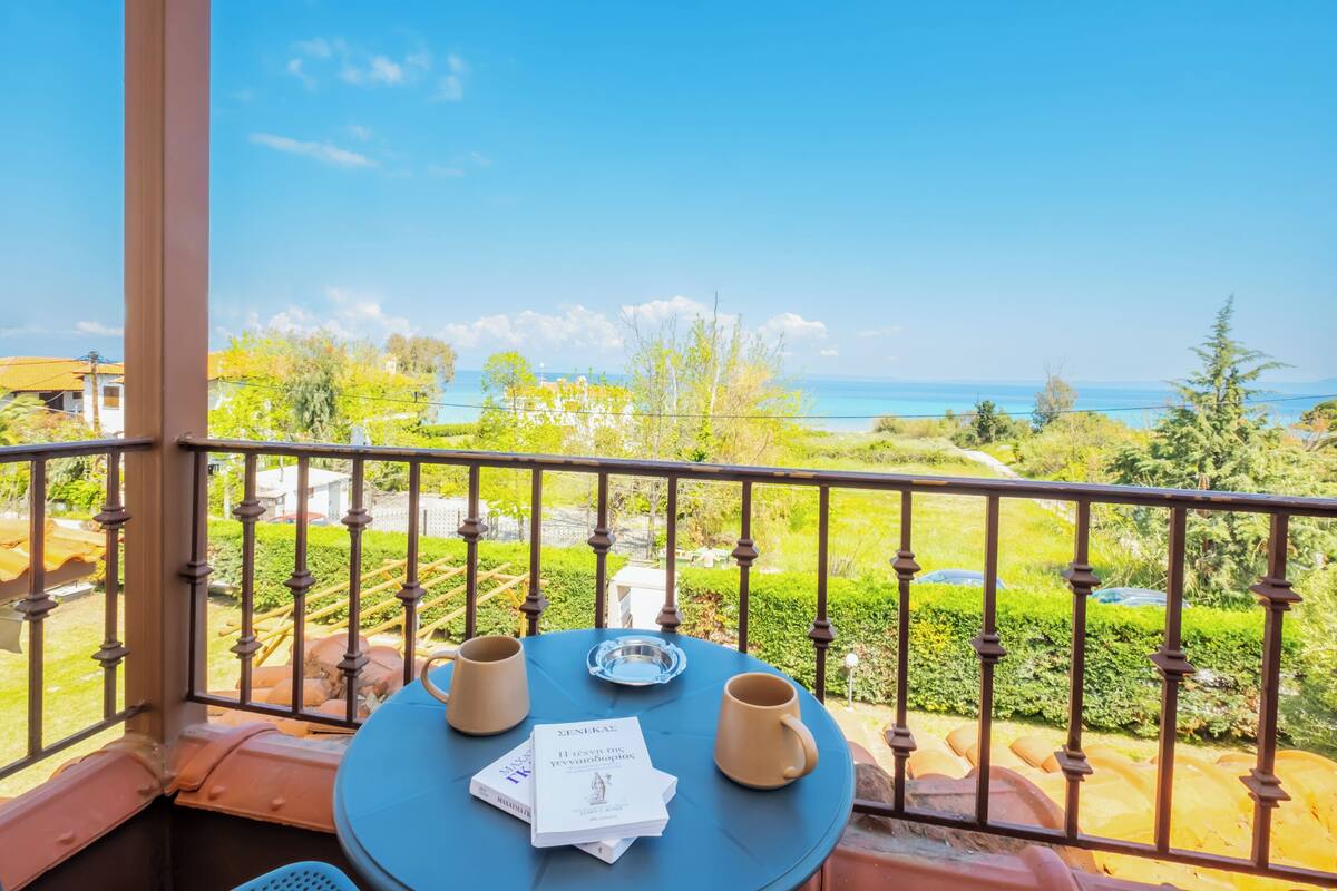 Varanda com mesa, duas canecas e papéis, vista para paisagem costeira com vegetação e edifícios.