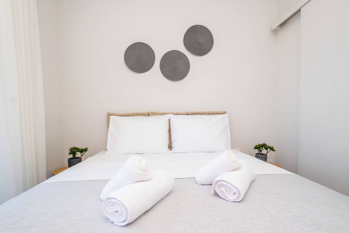 Cama bem arrumada com lençóis brancos e toalhas enroladas, duas pequenas plantas em vasos e três decorações circulares na parede.