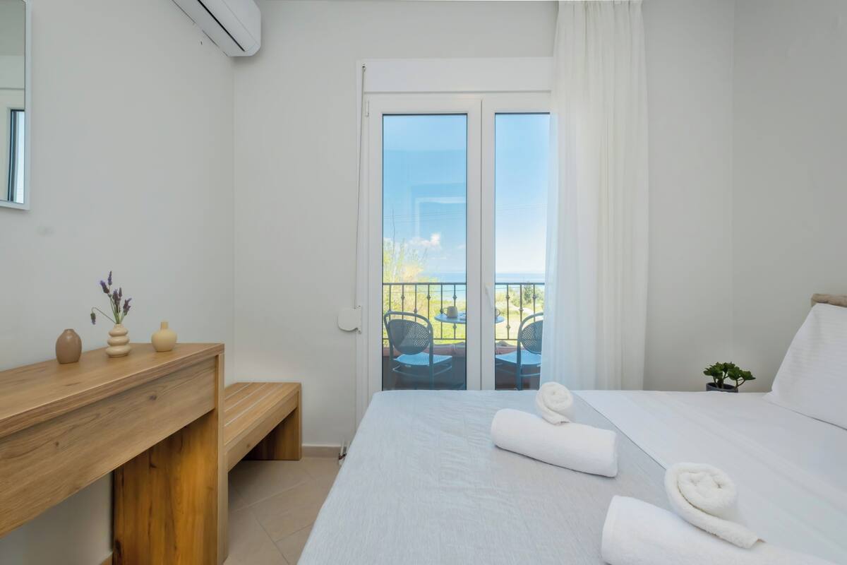 Quarto de hotel com uma cama, um banco de madeira, uma mesinha com vasos, uma varanda com cadeiras e um vaso de planta.