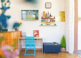 Uma área de recepção com uma mesa de madeira, uma cadeira azul, um pequeno banquinho azul, um relógio, uma prateleira com livros e garrafas, e uma moldura com uma fotografia.