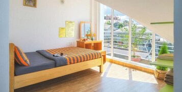 Quarto com cama, escrivaninha, janela e vista para prédios, e uma planta.