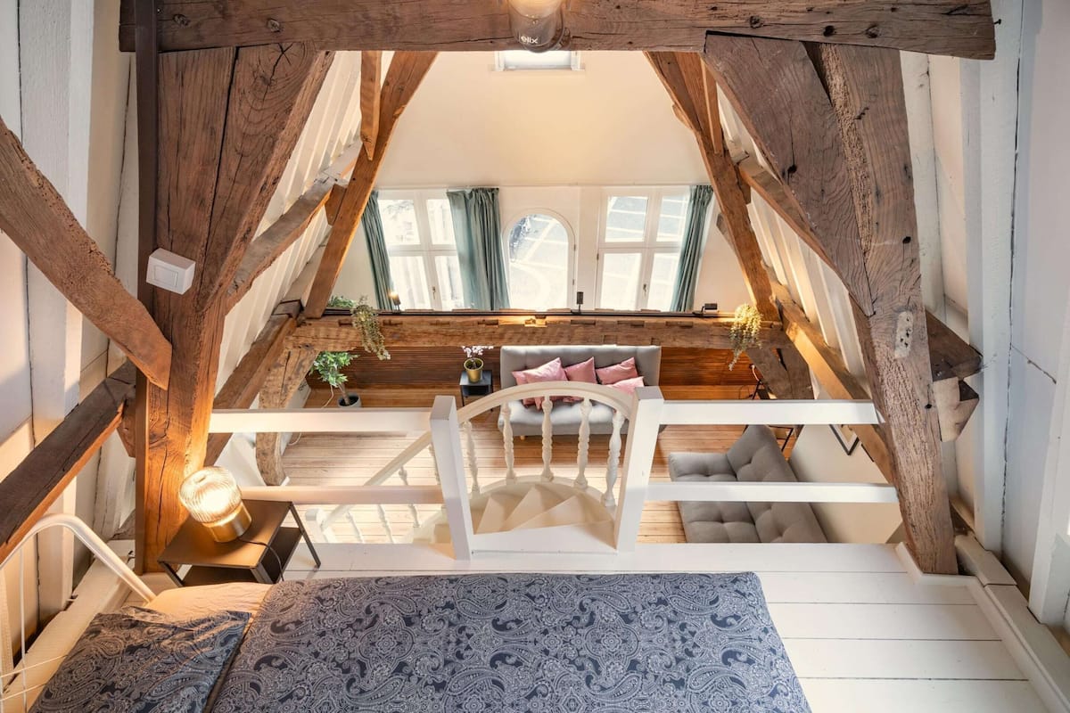Quarto em estilo loft com escada de madeira, cama branca e vista para a sala de estar.