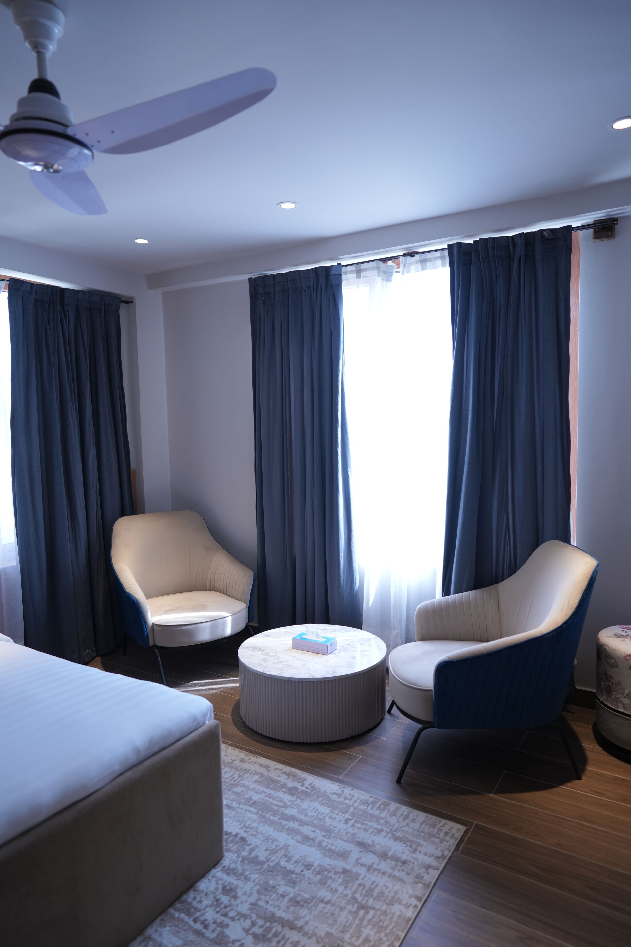 Quarto de hotel moderno com cama, duas poltronas, uma mesa de centro redonda e um ventilador de teto.