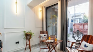 Uma varanda com mesa de madeira, cadeiras, um vaso de planta e vista para um edifício e uma cerca vermelha.