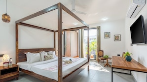 Uma cama de dossel com estrutura de madeira, uma mesa de cabeceira com abajur, uma escrivaninha de madeira e uma TV fixada na parede.