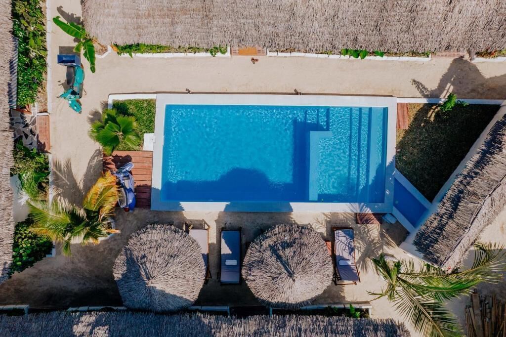 Vista aérea de uma piscina retangular com um deck ao redor, cadeiras de espreguiçadeira e guarda-sóis de palha.