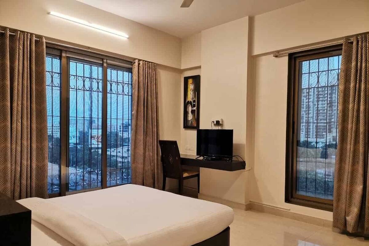 Quarto de hotel com uma cama, uma escrivaninha com computador e janelas amplas com cortinas.