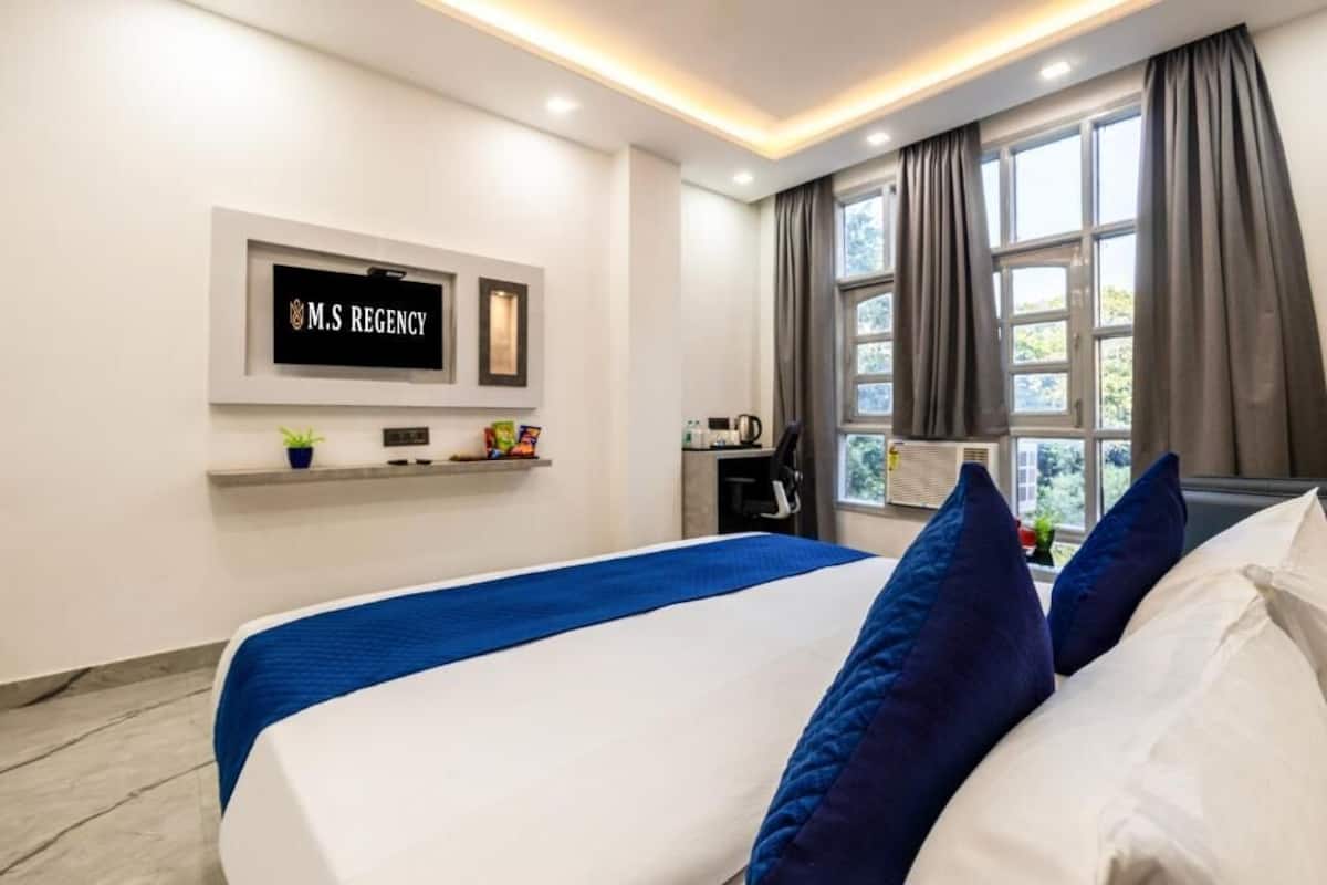Quarto de hotel com uma cama, televisão fixada na parede, janela com cortinas e uma pequena prateleira com objetos.
