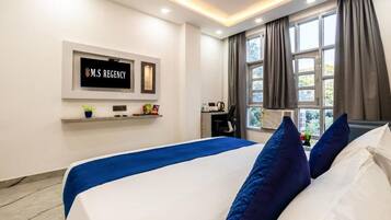 Quarto de hotel com uma cama, televisão fixada na parede, janela com cortinas e uma pequena prateleira com objetos.