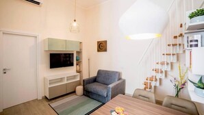 Uma sala de estar moderna com uma televisão de tela plana, um sofá cinza, uma mesa de jantar com cadeiras e uma luminária pendente decorativa.