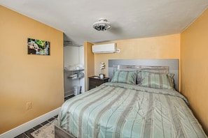 Quarto com cama, ar-condicionado, ventilador de teto, mesa de cabeceira, pequena mesa com abajur e um quadro na parede.