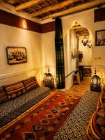 Interior tradicional marroquino com teto de madeira, tapete estampado, sofá com almofadas e lanternas fixadas na parede.